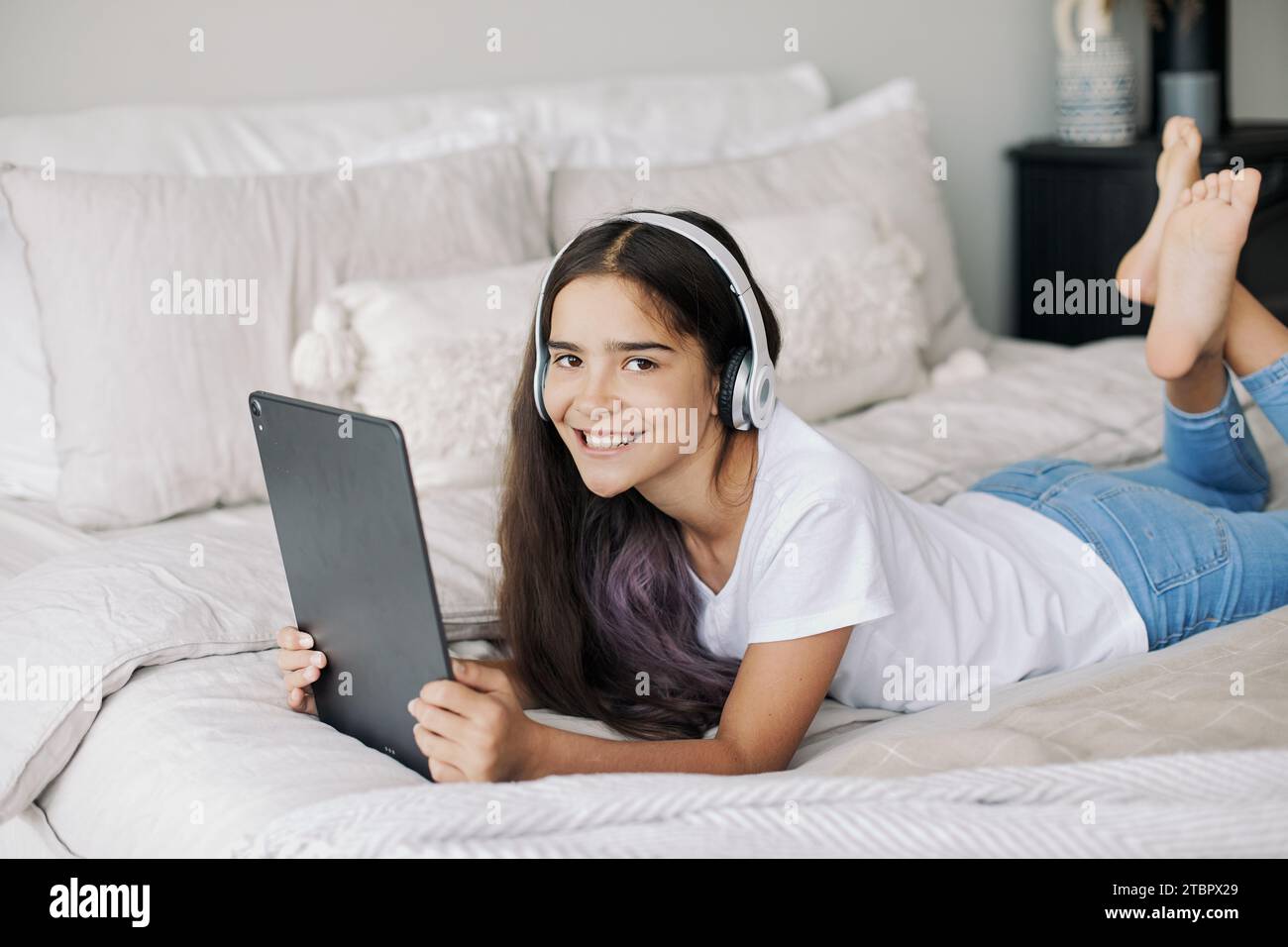 Adorable pre-teen girl in wireless headphones lying on bed with digital tablet device. Young ...