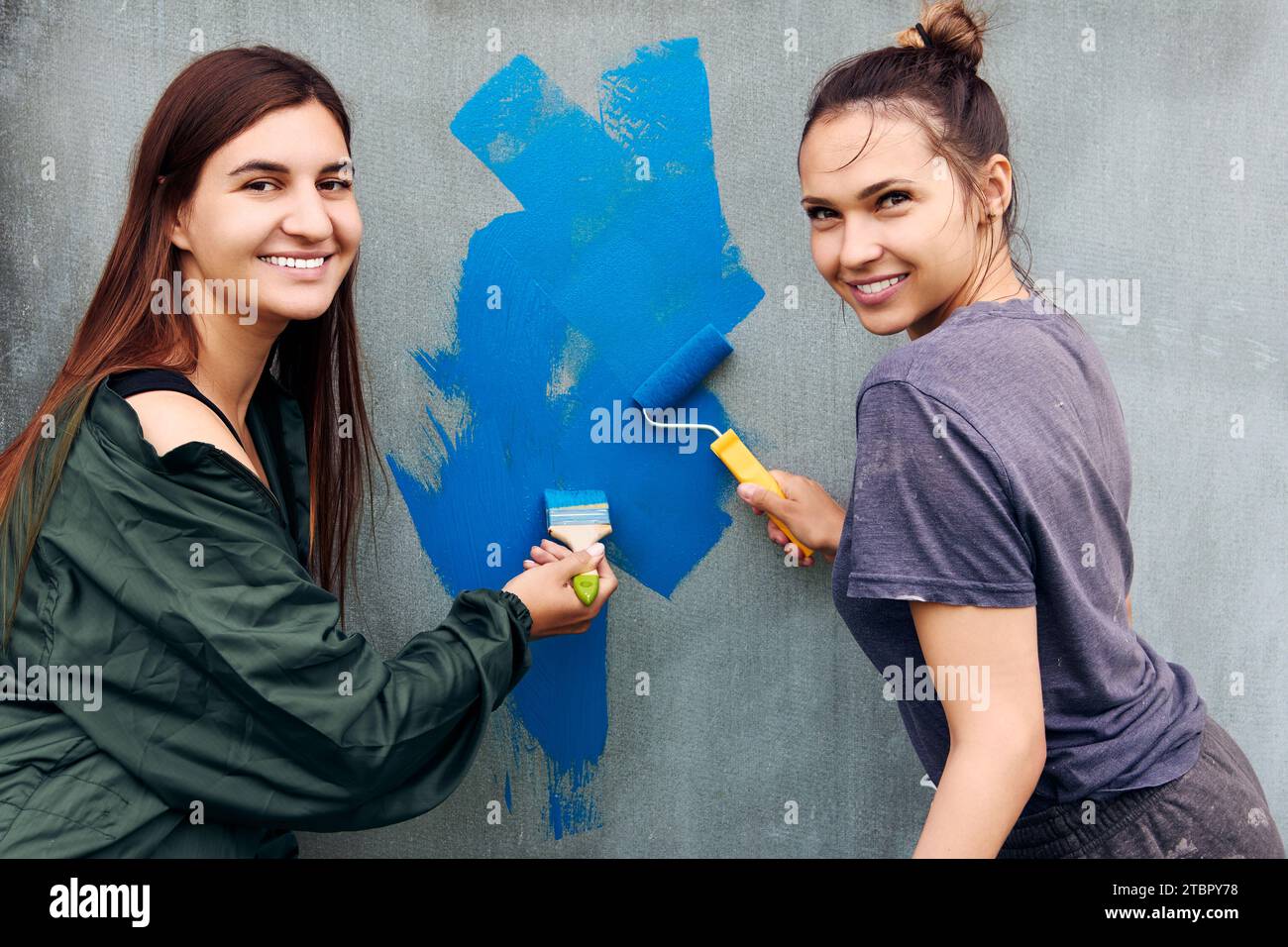 Using blue paint to painting gray wall, smiling young women working as ...