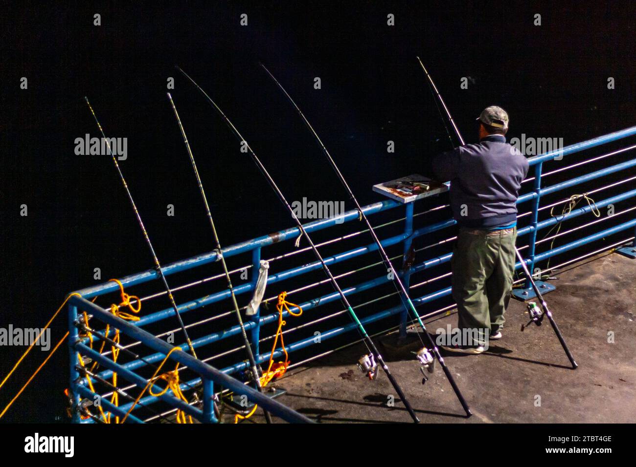 A fisherman fishing off a pier at night with multiple fishing rods ...