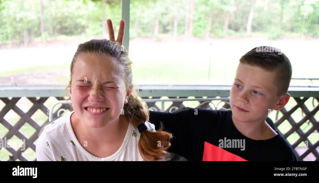 Young pre-teen grimaces as her kid brother does rabbit ears behind her ...