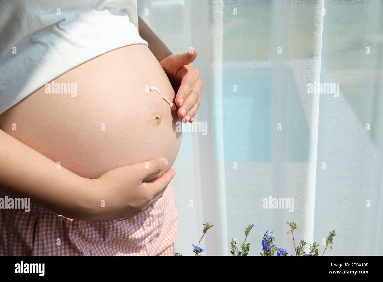 Pregnant woman applying stretch mark cream on her belly by the window ...