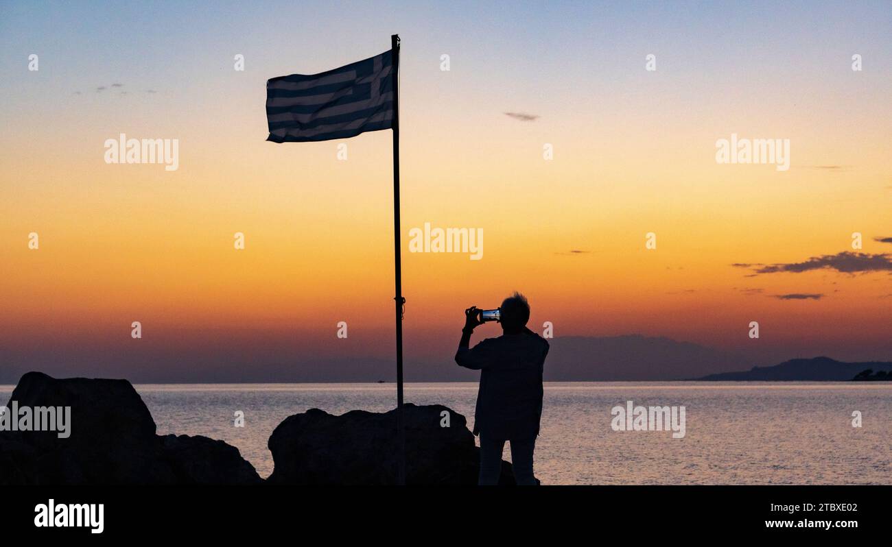 A lady stands on the shore and takes photographs of the Greek flag that ...