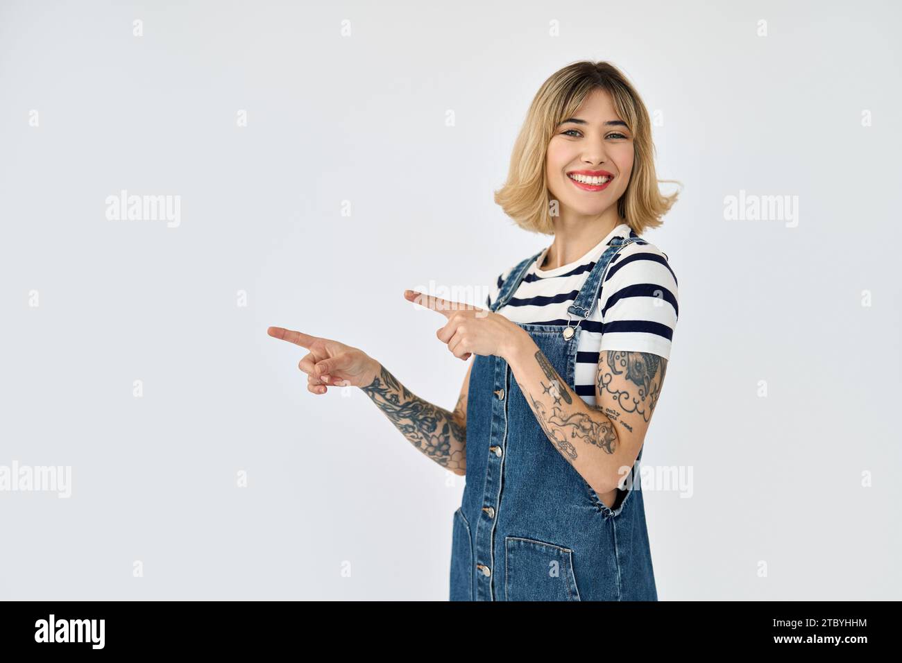 Happy gen z blonde young woman with tattoos pointing aside isolated on ...