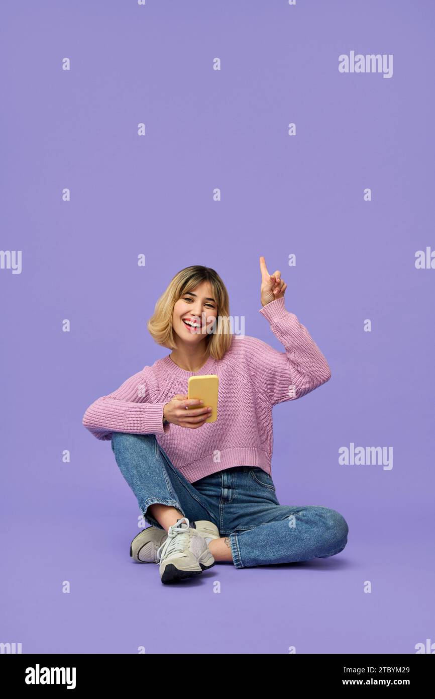 Happy pretty gen z blonde girl using mobile phone pointing up isolated ...