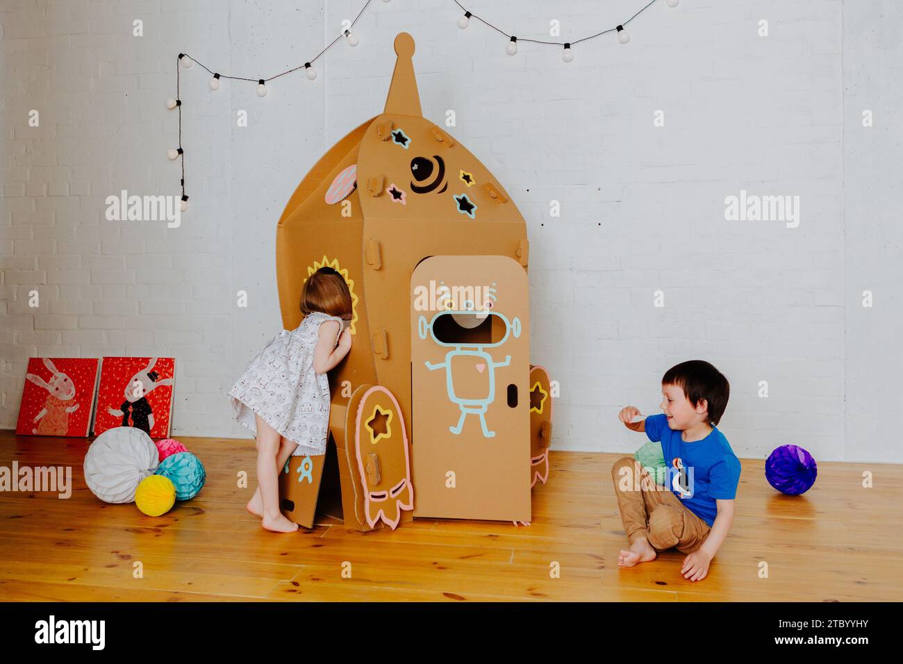 Joyful siblings running after each other and around cardboard rocket ...