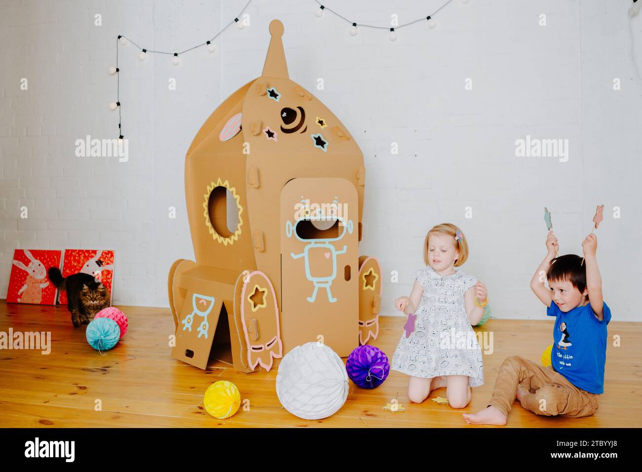 Joyful siblings running after each other and around cardboard rocket ...