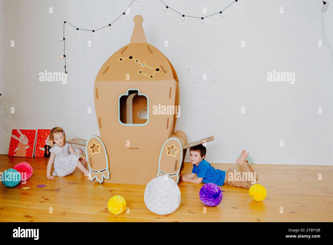 Joyful siblings running after each other and around cardboard rocket ...