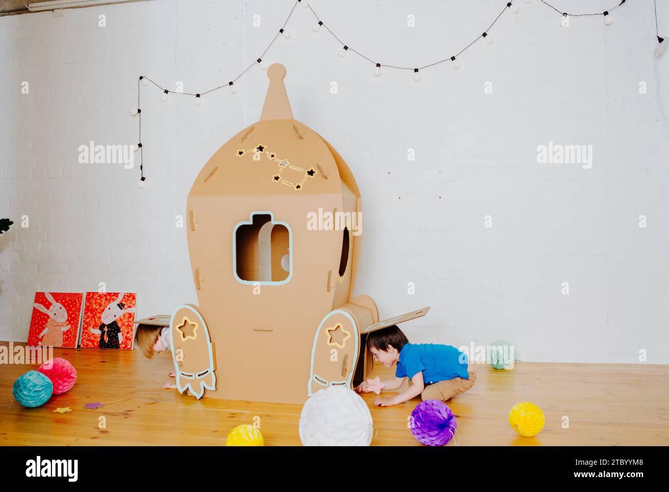 Joyful siblings running after each other and around cardboard rocket ...