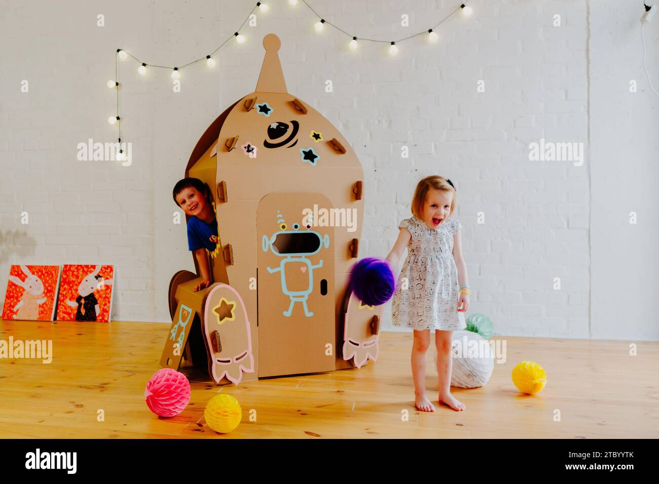 Joyful siblings running after each other and around cardboard rocket ...