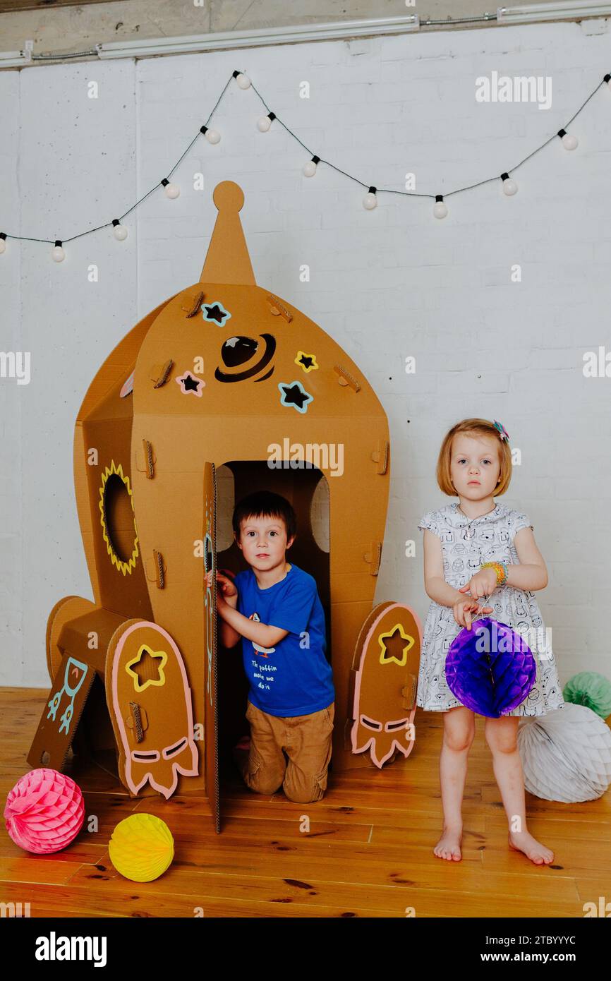 Joyful siblings running after each other and around cardboard rocket ...
