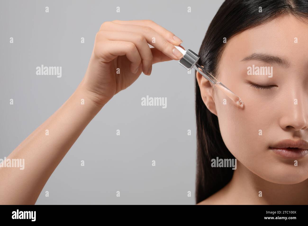 Beautiful young woman applying cosmetic serum onto her face on grey ...