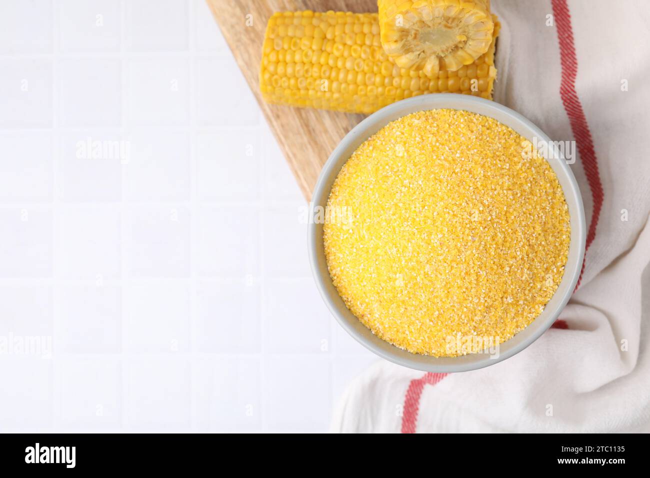 Raw cornmeal in bowl and corn cobs on white tiled table, top view ...