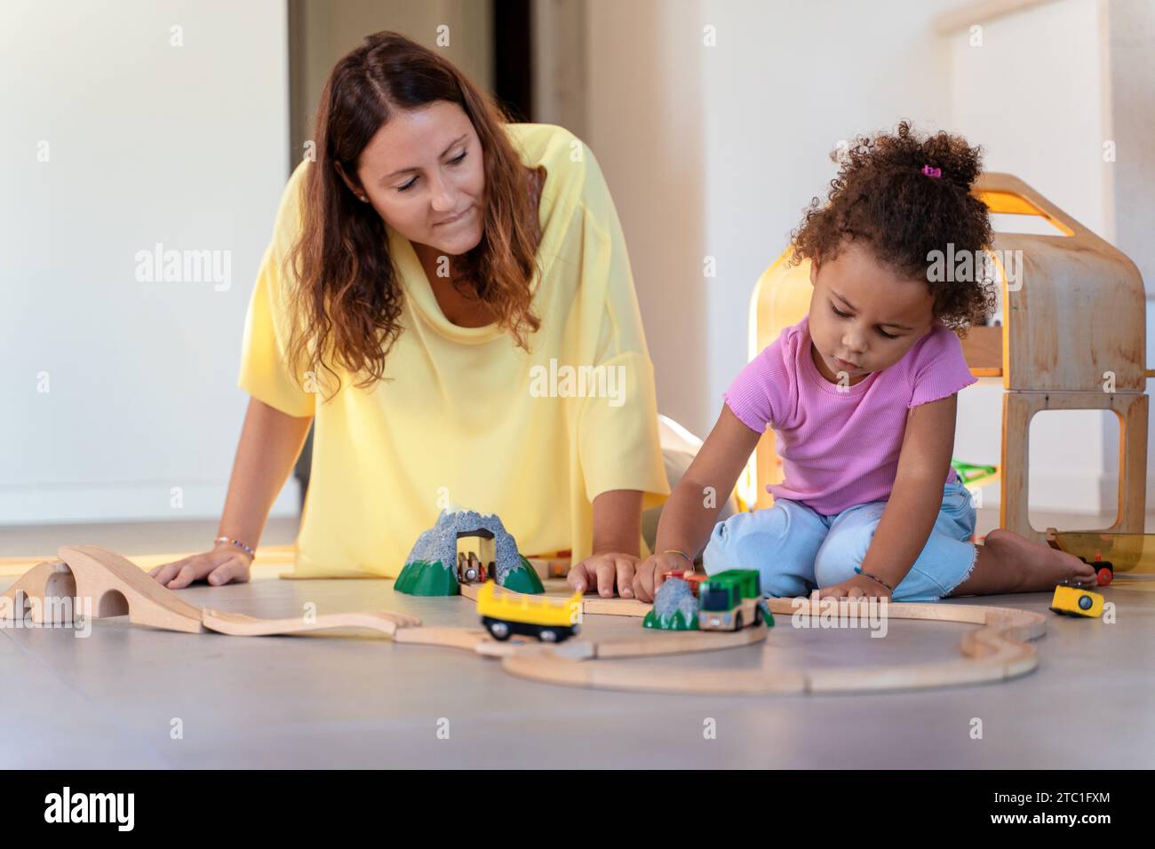 Interracial mother and daughter focus on building a wooden train track, enjoying creative ...