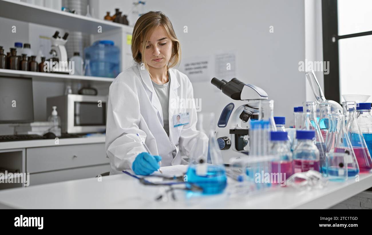 Dazzling young blonde scientist in busy lab scribbles down document of ...