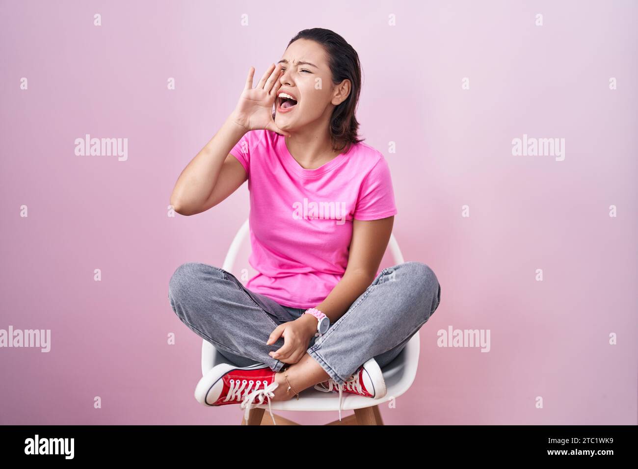 Hispanic young woman sitting on chair over pink background shouting and ...