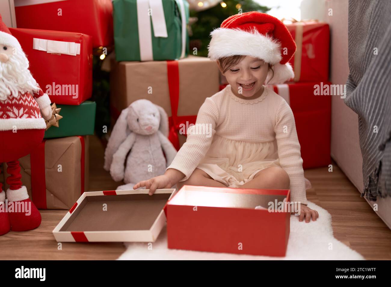 Adorable girl unpacking gift sitting by christmas tree at home Stock ...