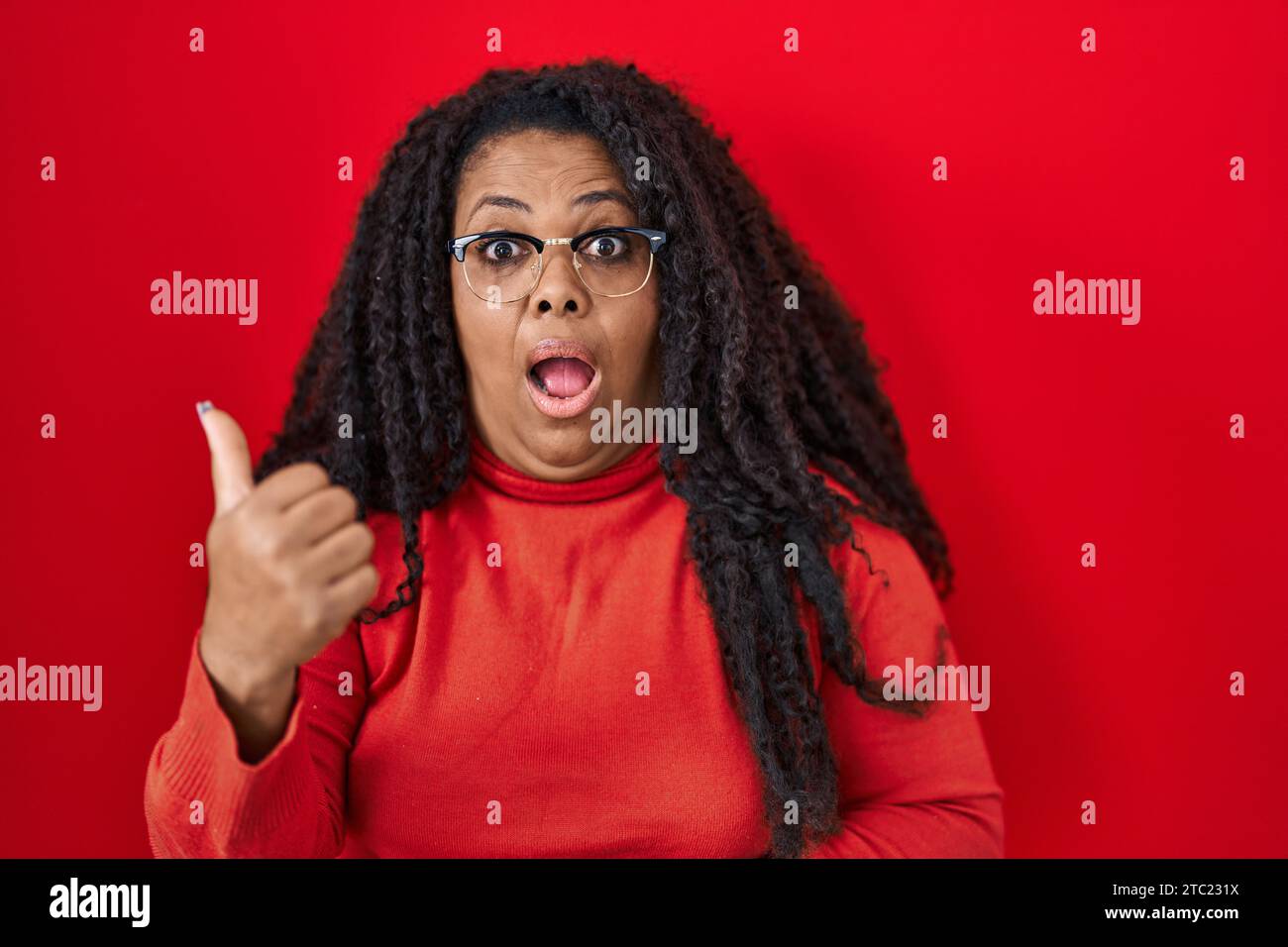 Plus size hispanic woman standing over red background surprised ...