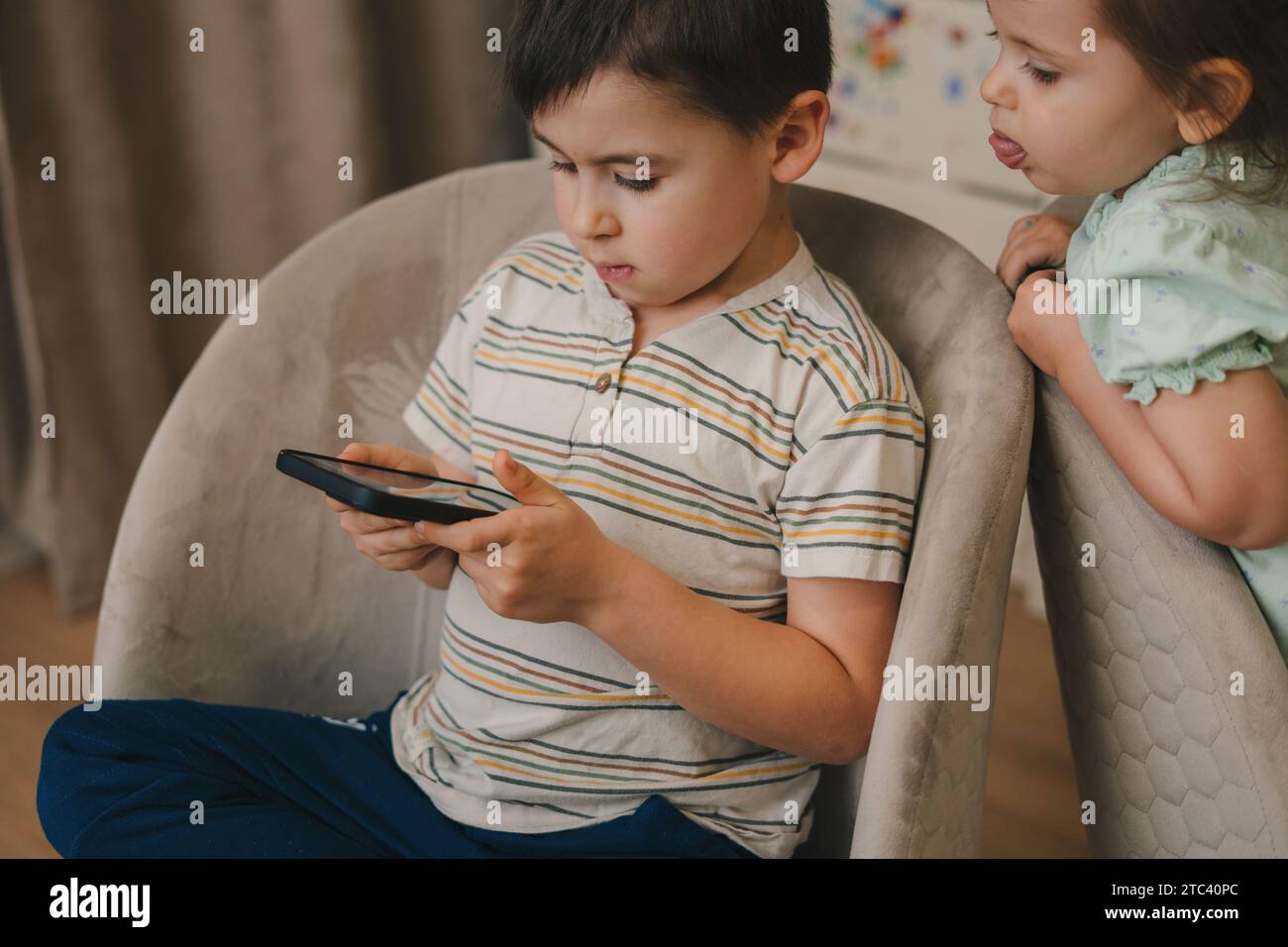 African Brother And Sister Sitting On Armchairs At Home Using Mobile Phone Looking At Device