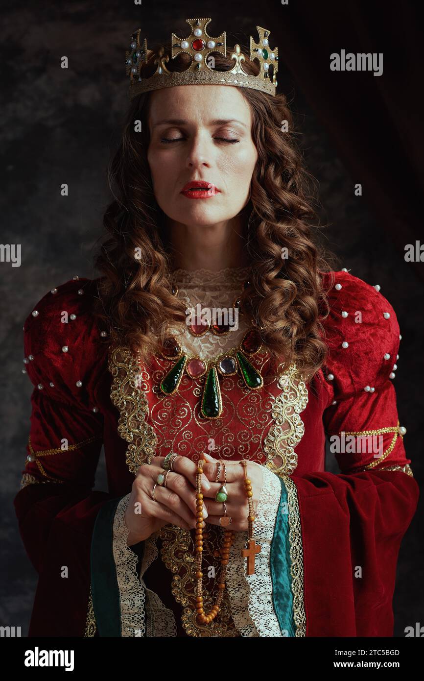 medieval queen in red dress with rosary and crown praying on dark gray ...