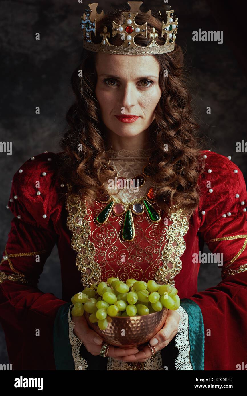 medieval queen in red dress with plate of grapes and crown on dark gray ...