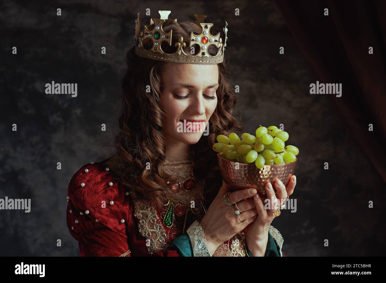 smiling medieval queen in red dress with plate of grapes and crown on ...