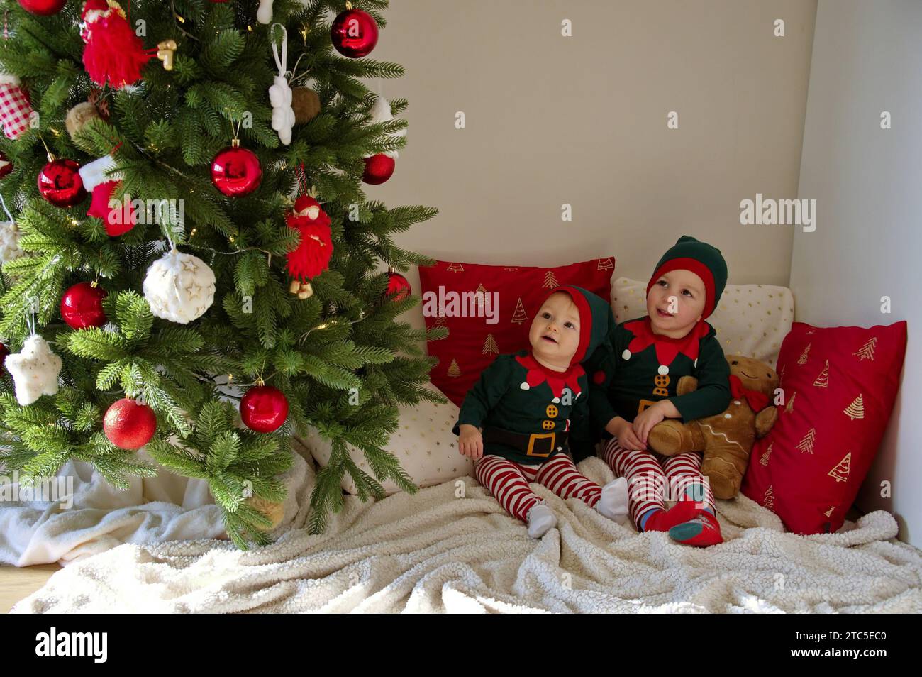 Baby girl and toddler dressed as elf sitting in front of Christmas tree ...
