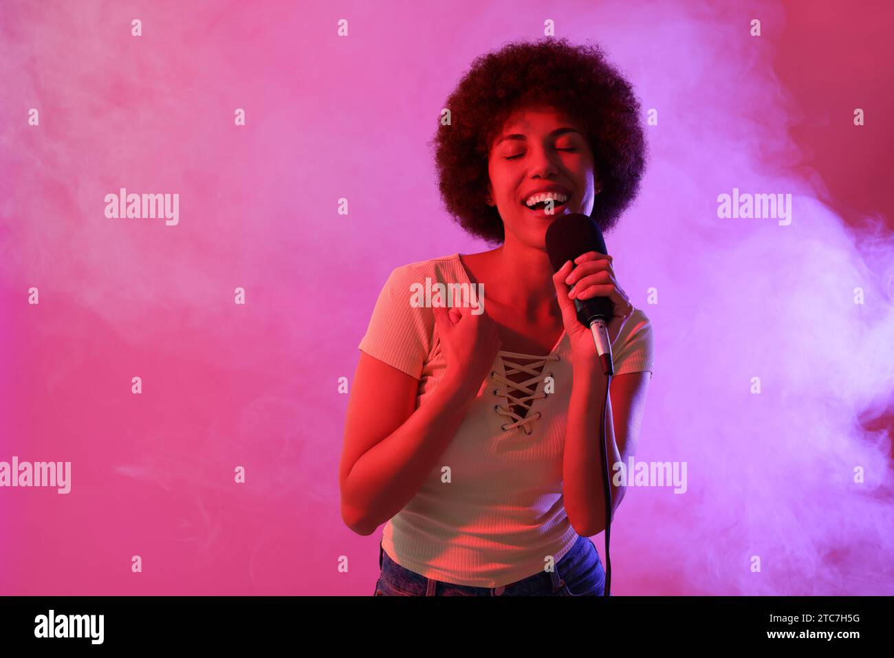 Curly young woman with microphone singing on pink background, space for ...
