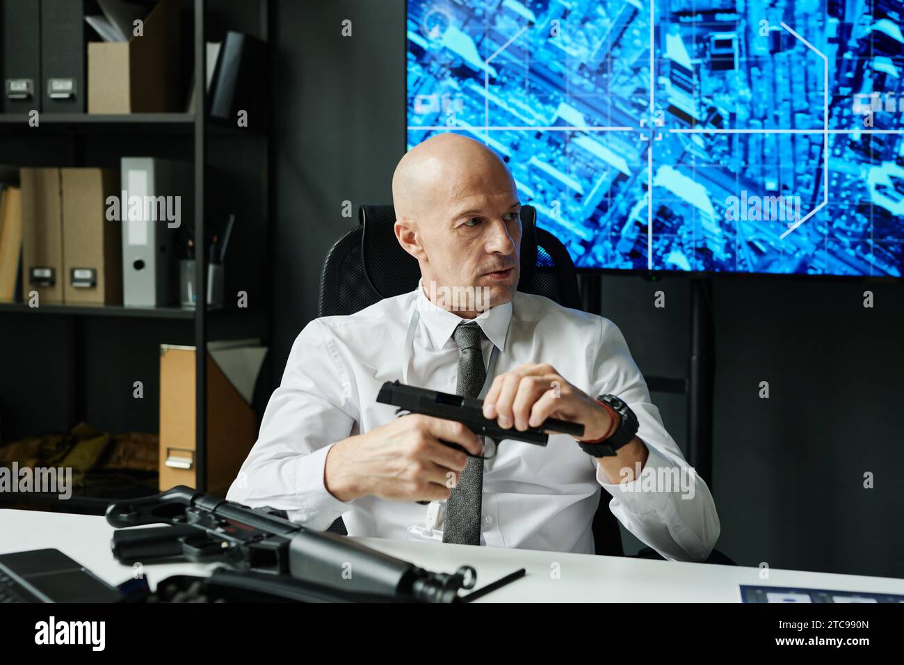 Bald mature man with pistol sitting by workplace during presentation of ...