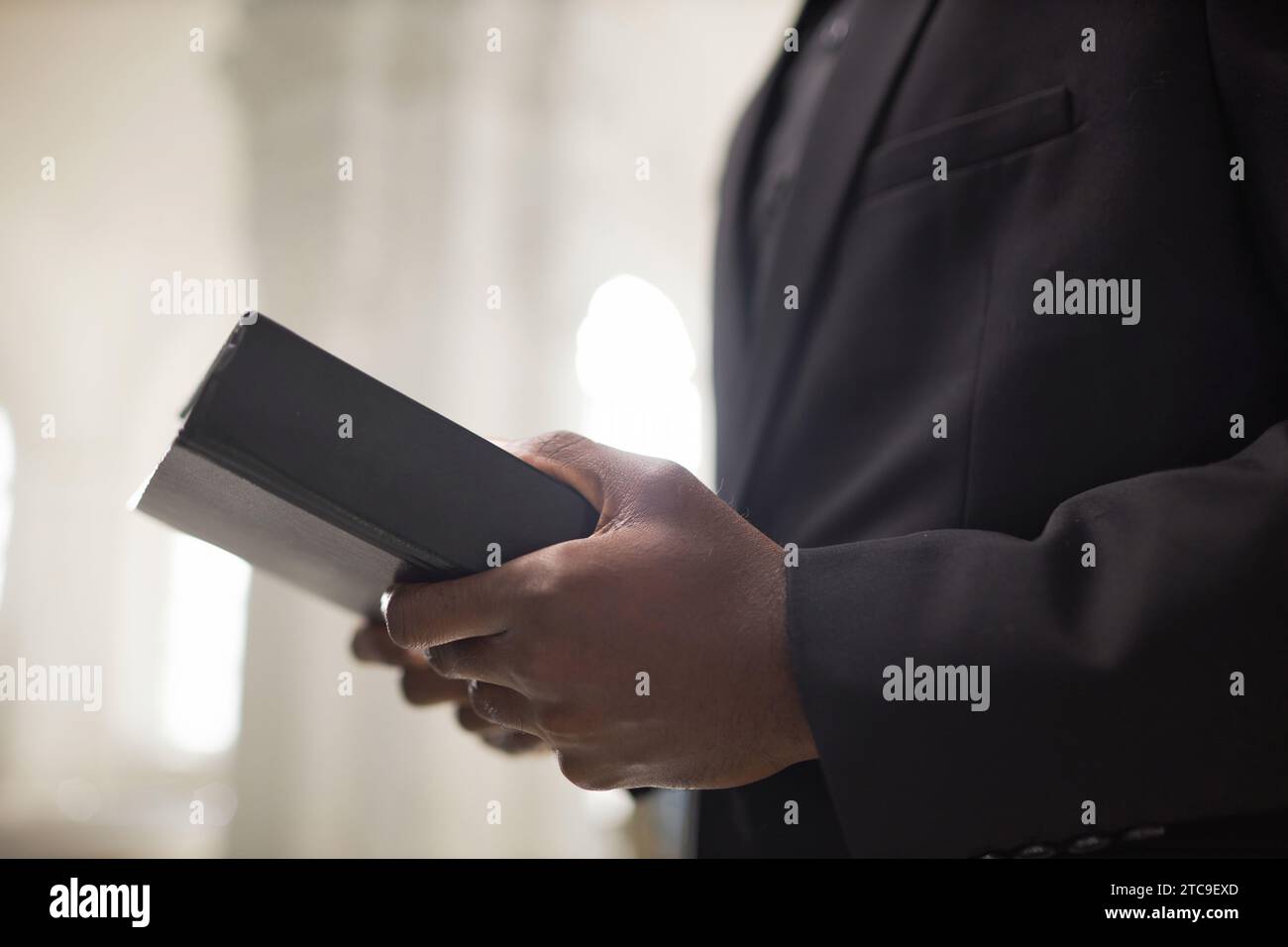 Side view closeup of unrecognizable Black priest holding Bible in hands ...