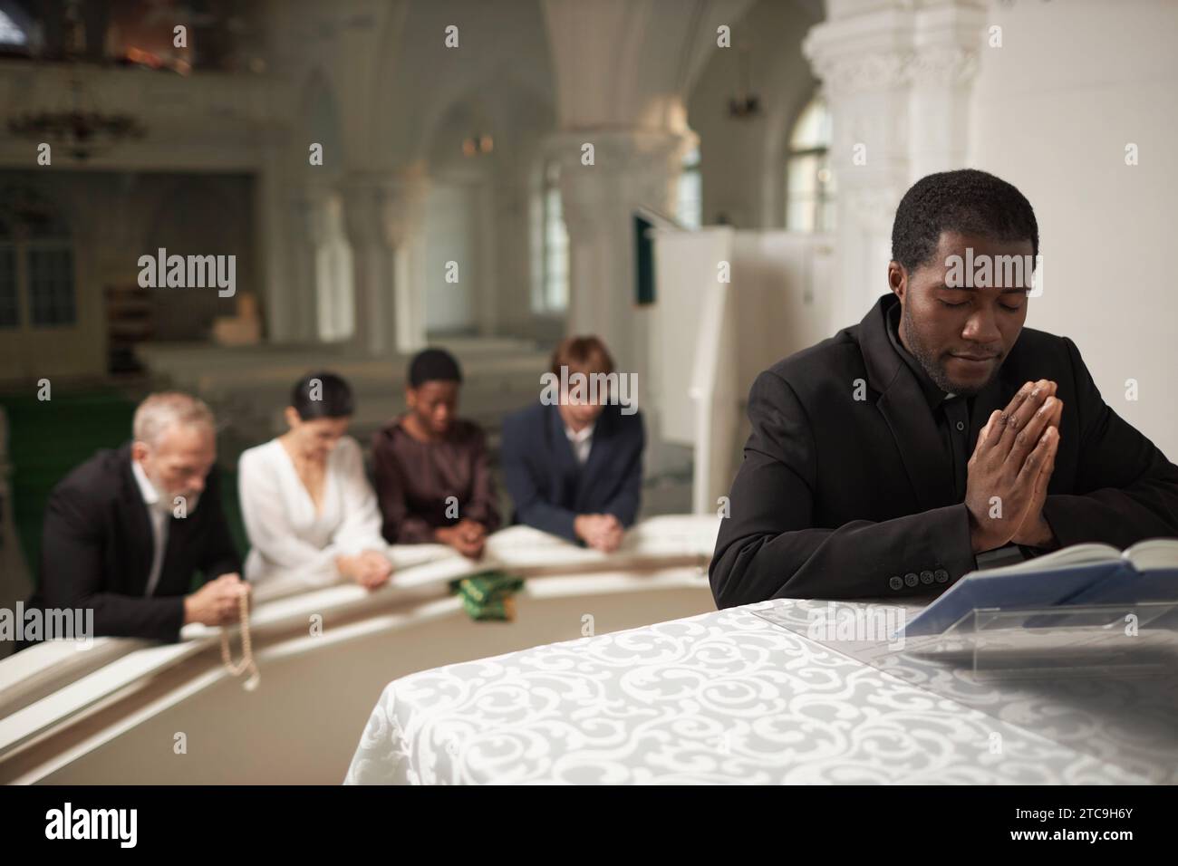 Portrait of young African American priest praying at altar with group ...