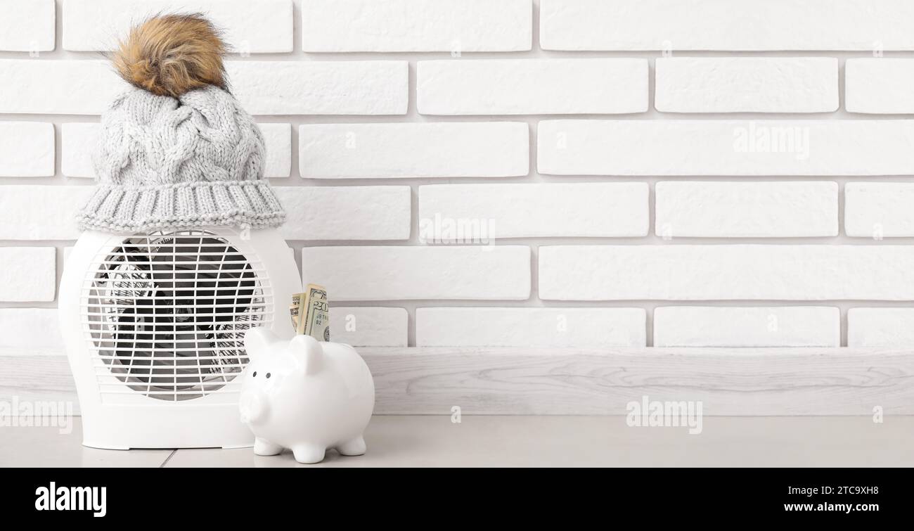 Electric radiator with knitted hat and piggy bank near white brick wall ...