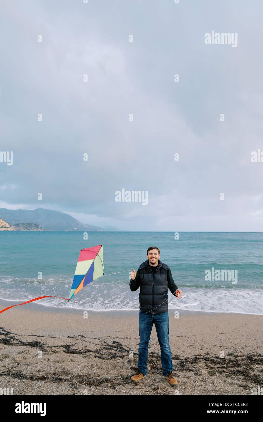 Young smiling man holding a colorful kite on a string on the beach ...