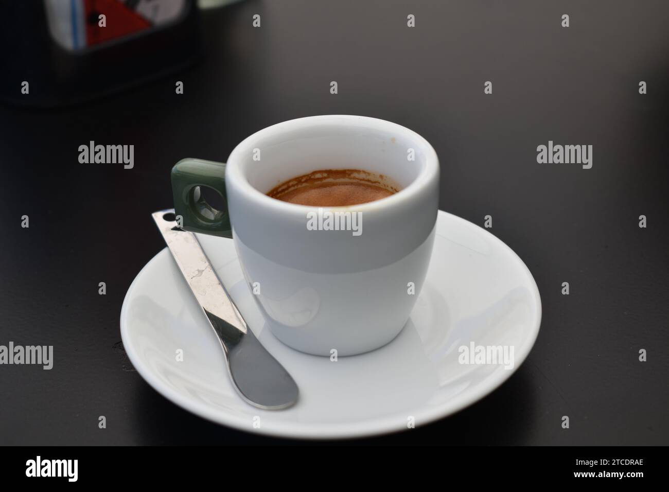 A cup of espresso on a small white plate with a small thin stainless ...