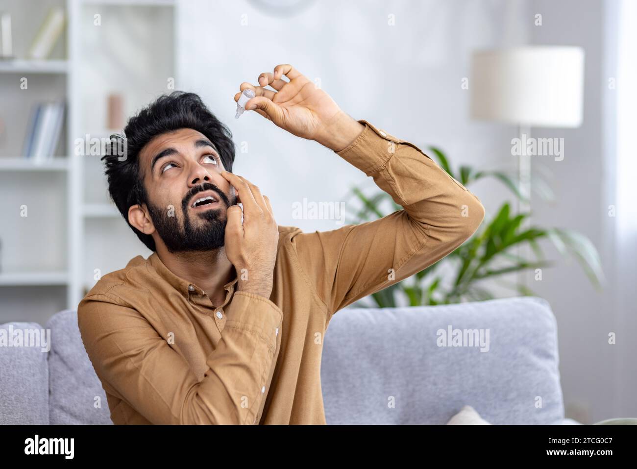 A young Indian man sits at home sick on the sofa and drops medicine in ...