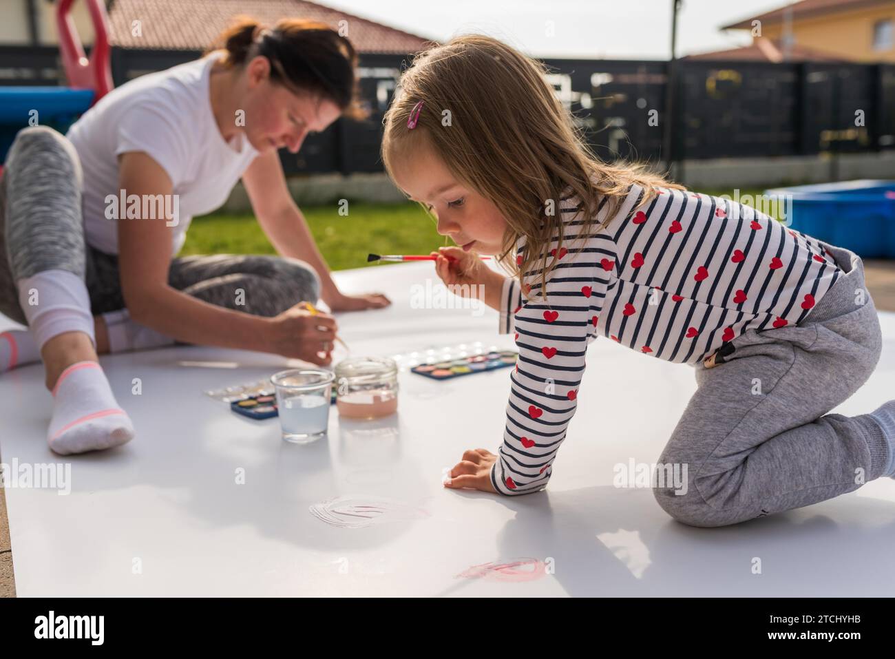 Focused Child with mother painting with poster paint outdoors on the ground on big sheet of ...
