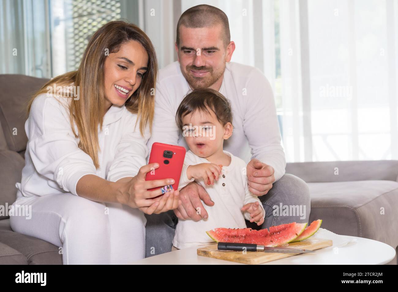 Family of three with a baby boy using mobile while eating fruit in the ...