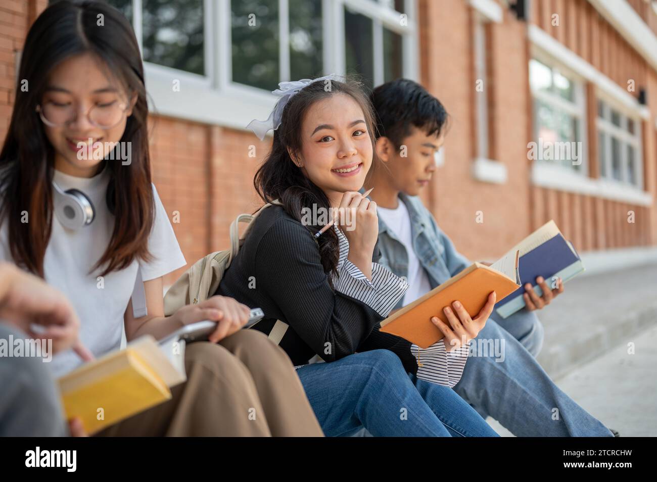 A positive, cute young Asian female college student sits on a footpath with her friends ...