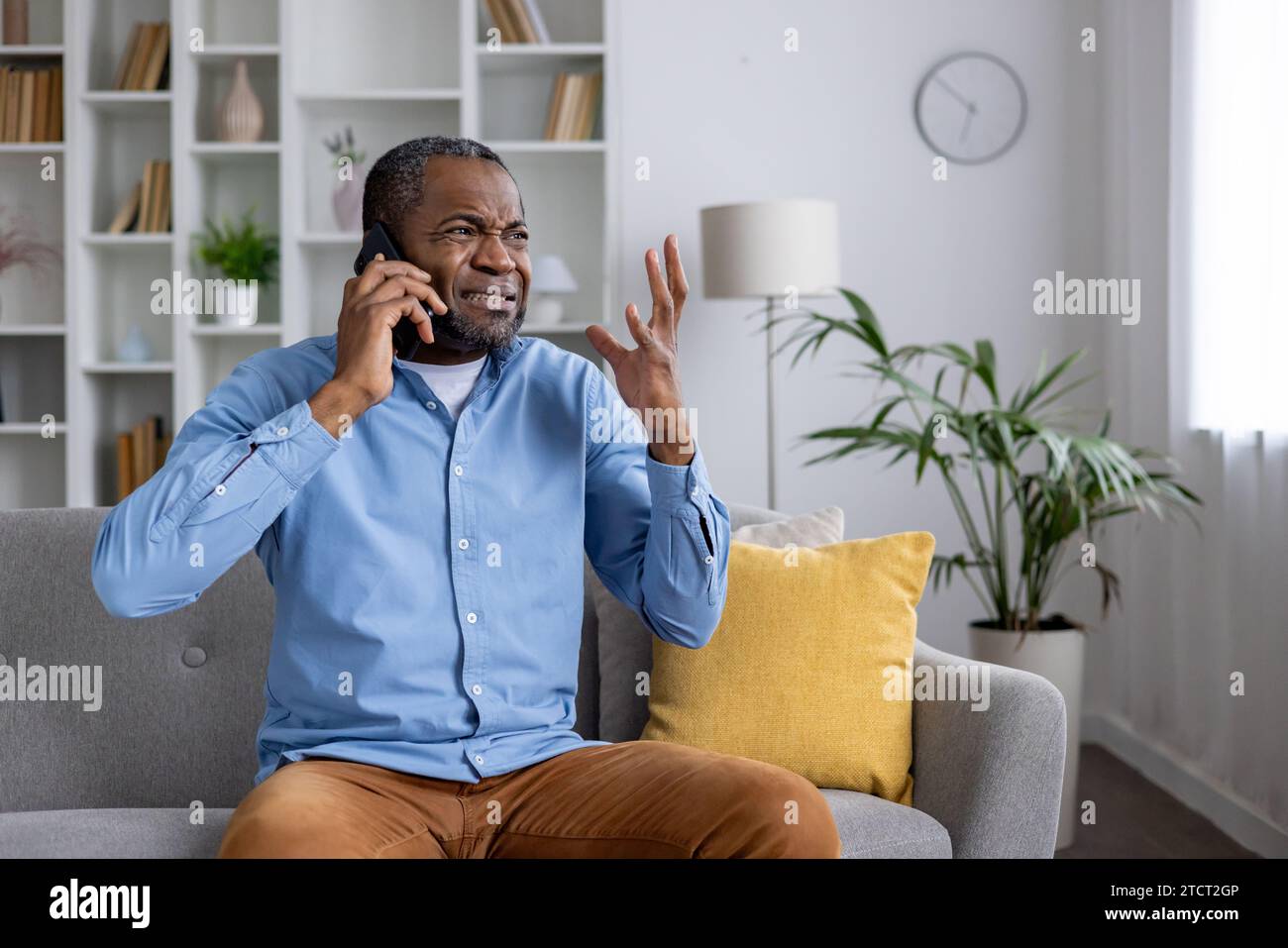 Angry nervous man sitting on sofa at home talking on the phone, unhappy ...