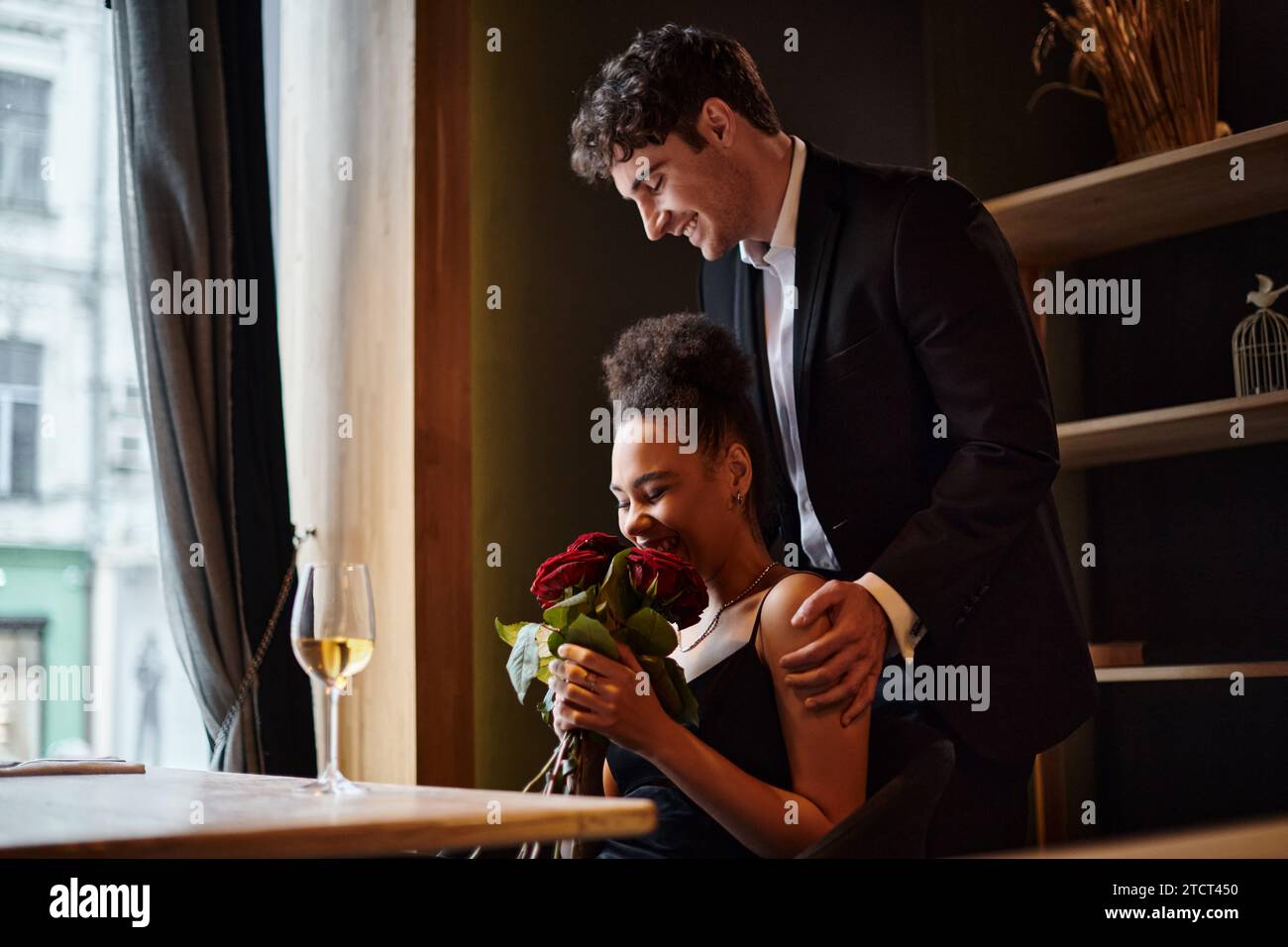 pleased african american woman smelling red roses near man in suit ...