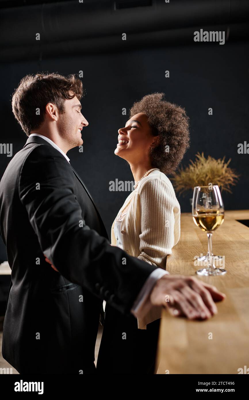 happy multicultural couple laughing while standing near bar counter ...