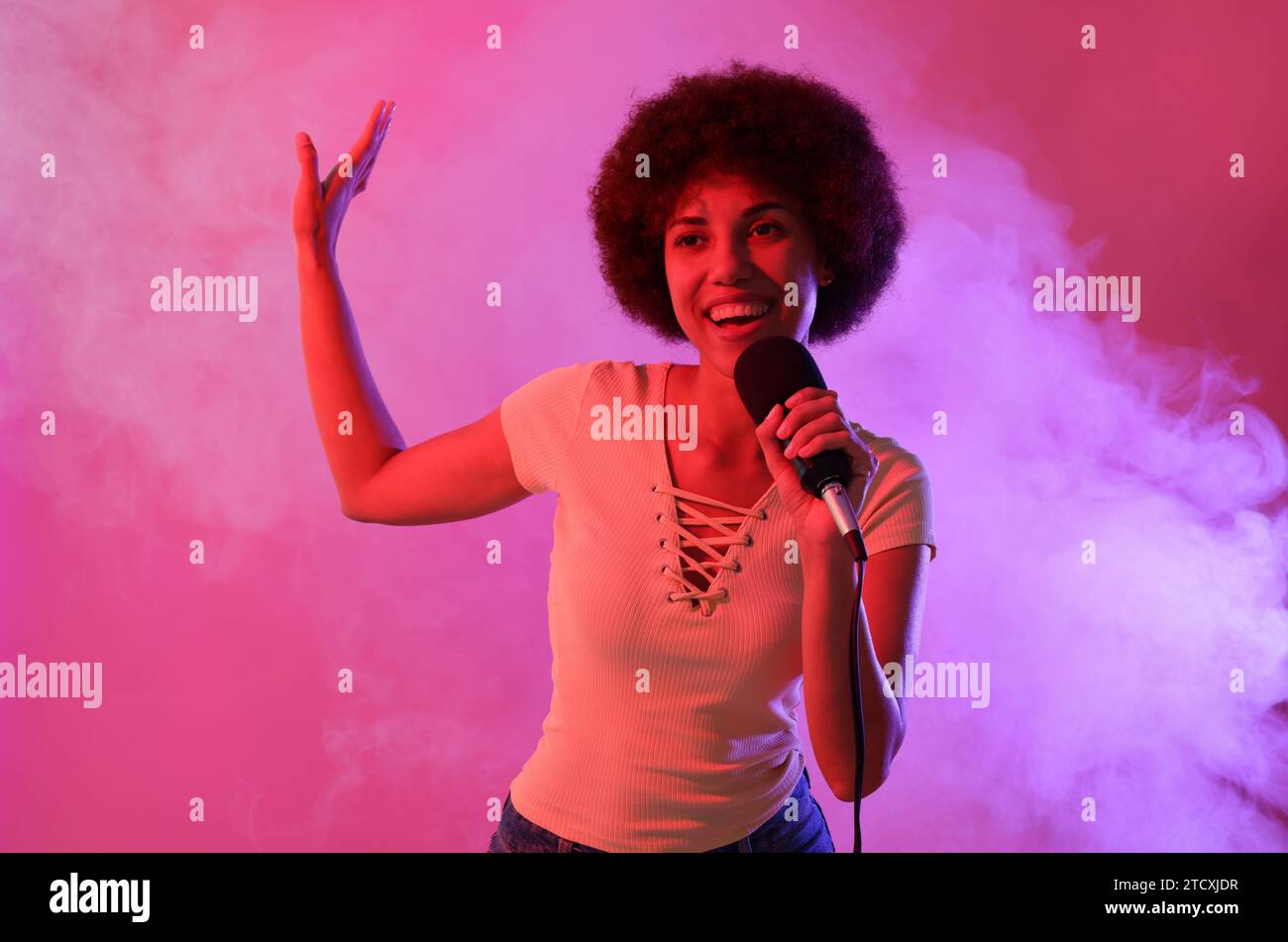 Curly young woman with microphone singing on pink background. Color ...