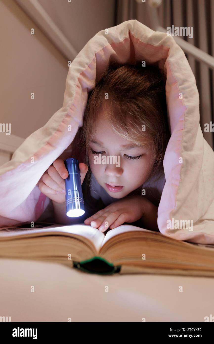 little girl lying in bed and reading book with flashlight under blanket ...