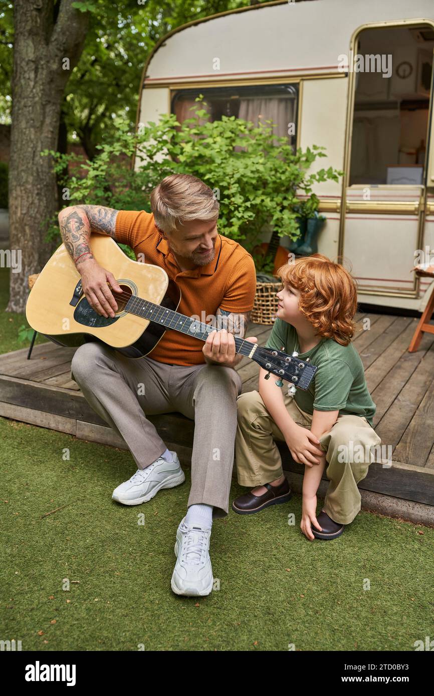 tattooed father playing acoustic guitar to smiling son next to trailer ...