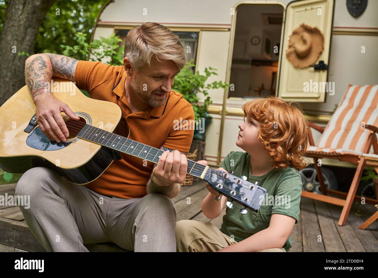 tattooed father playing acoustic guitar to attentive son next to ...