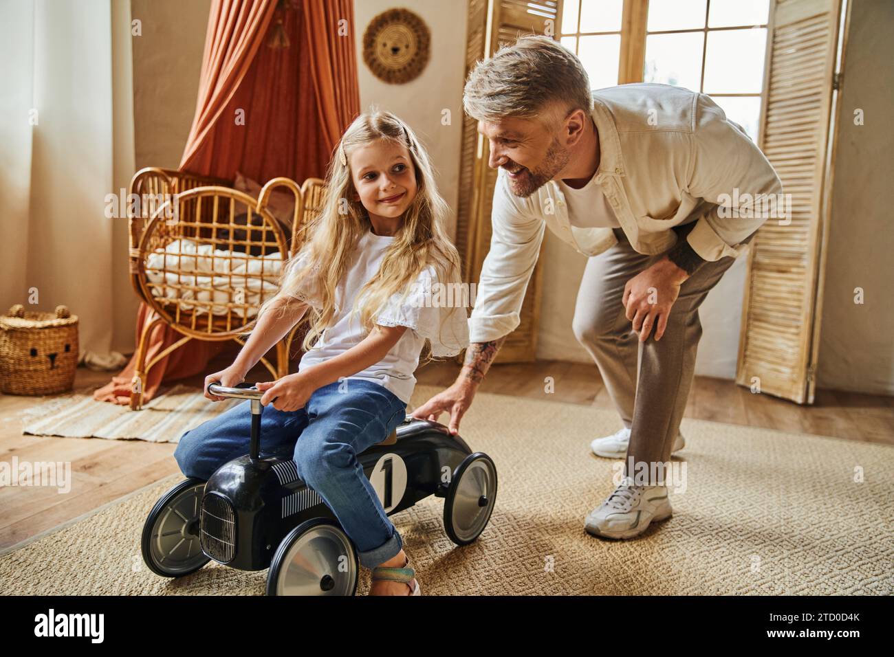 cheerful man assisting cute daughter riding toy car in modern living ...