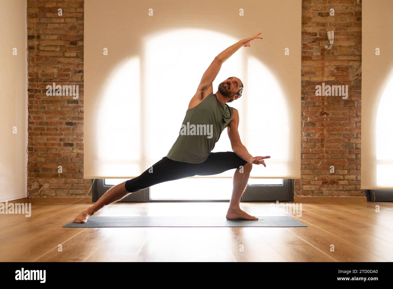 A man holds a yoga pose on a mat in a well-lit studio with brick walls ...
