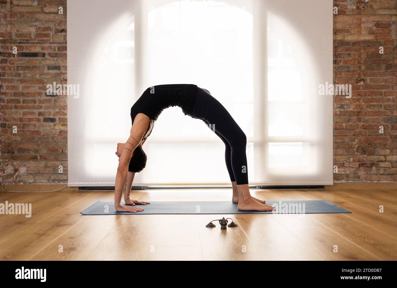 A woman is captured in a backbend yoga pose, the Wheel Pose, on her mat in a well-lit room with ...