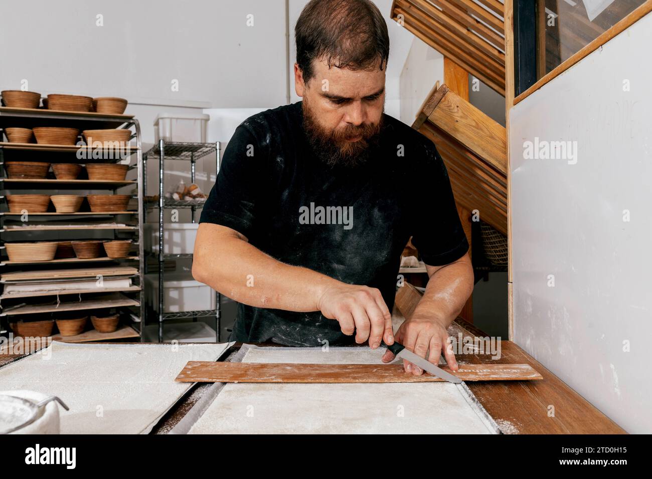 Serious male bearded baker with concentration cutting dough with wooden ...