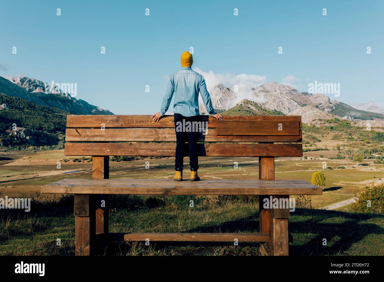 A man stands on a wooden big bench, facing a stunning mountain range ...
