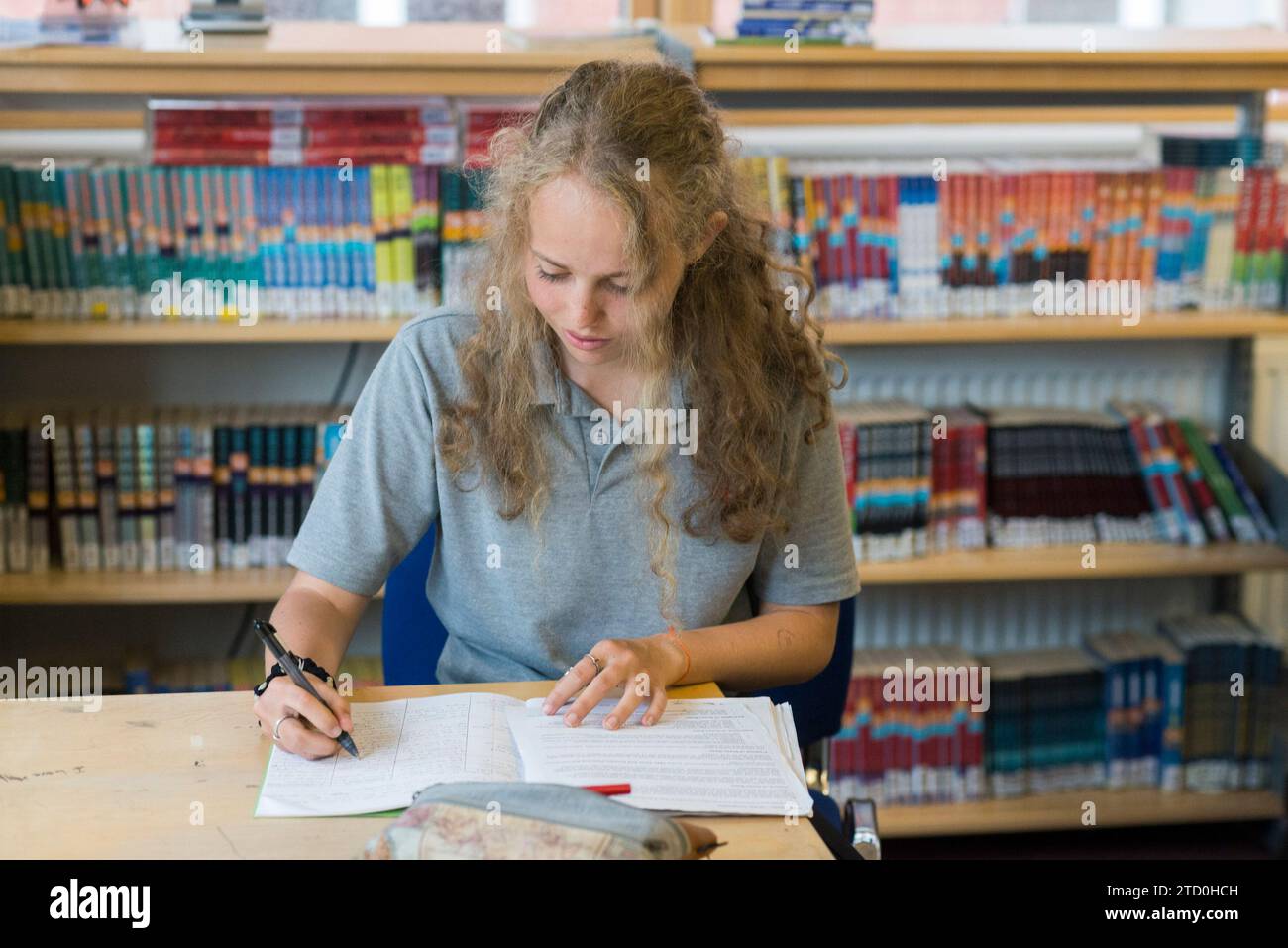 A female A level student sits at a desk in a school library revising ...
